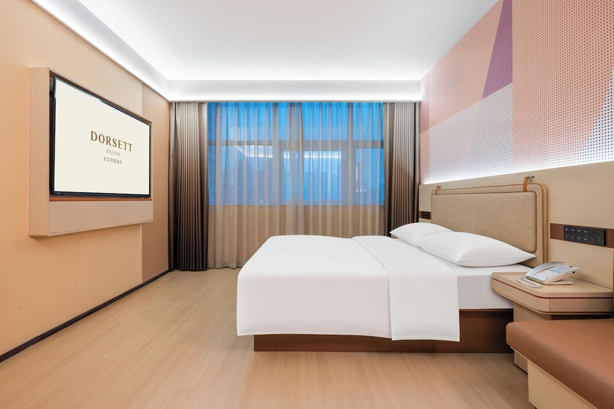 Quarto de hotel com uma cama, mesa de cabeceira com telefone, televisão fixada na parede e uma grande janela com cortinas.