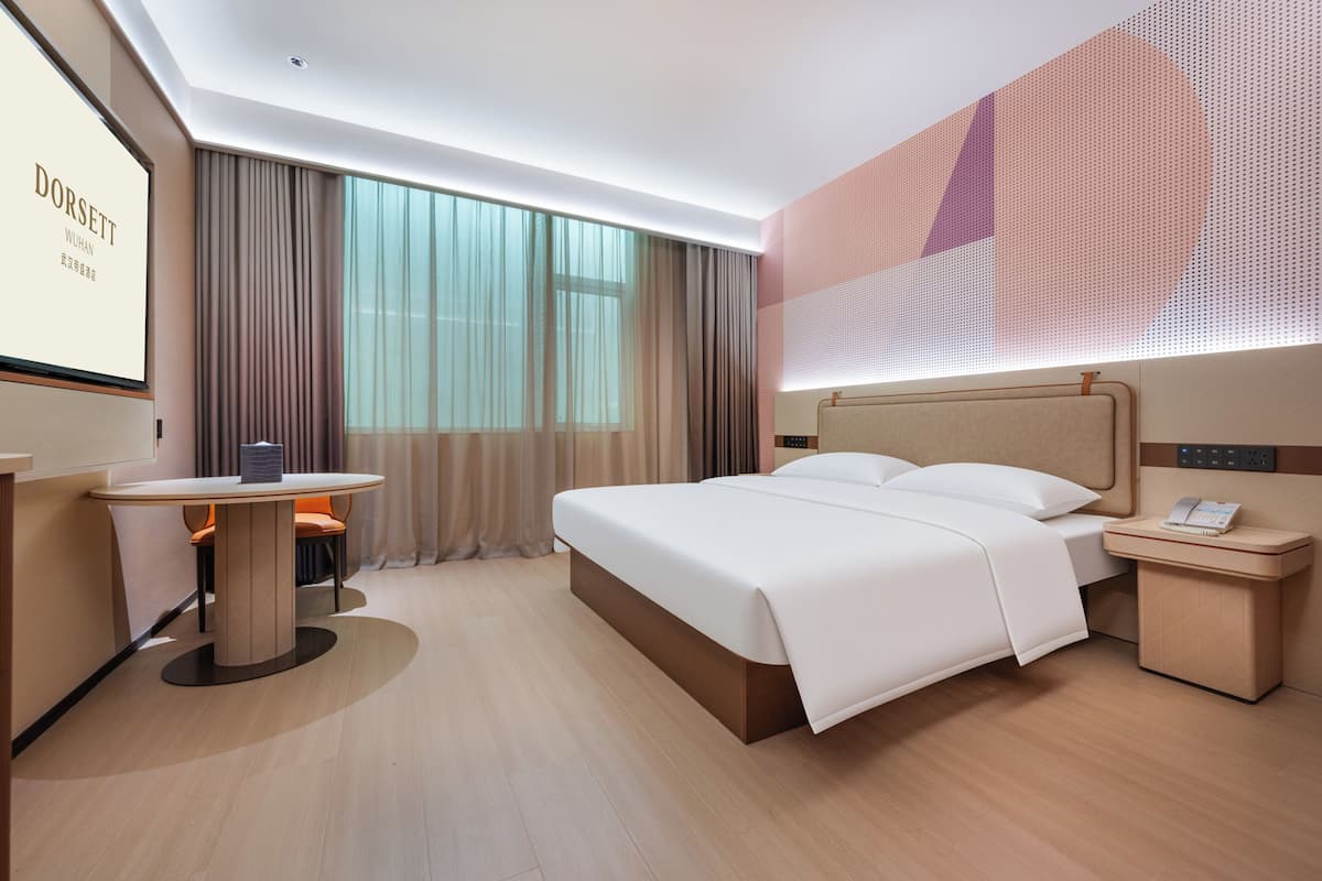 Quarto de hotel com cama, mesa de cabeceira, escrivaninha e cortinas.