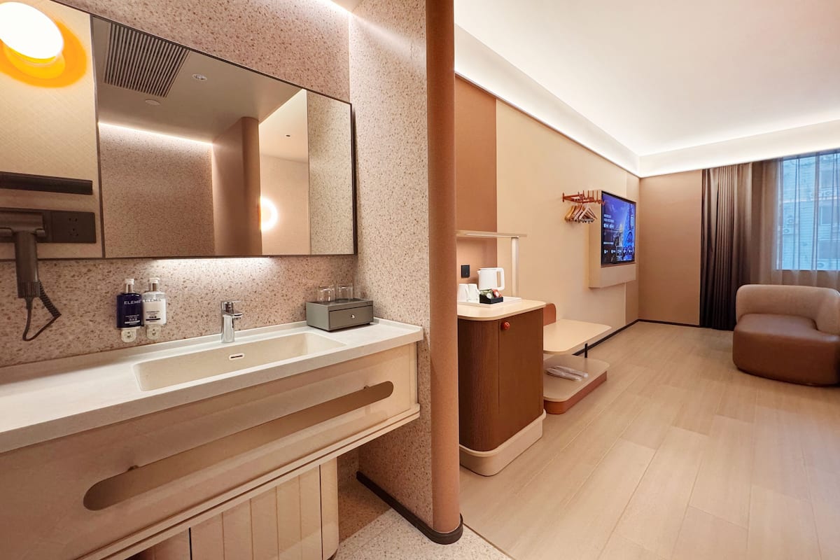 Quarto de hotel moderno com um espelho grande, pia, artigos de higiene, televisão de tela plana, área de estar e janela.