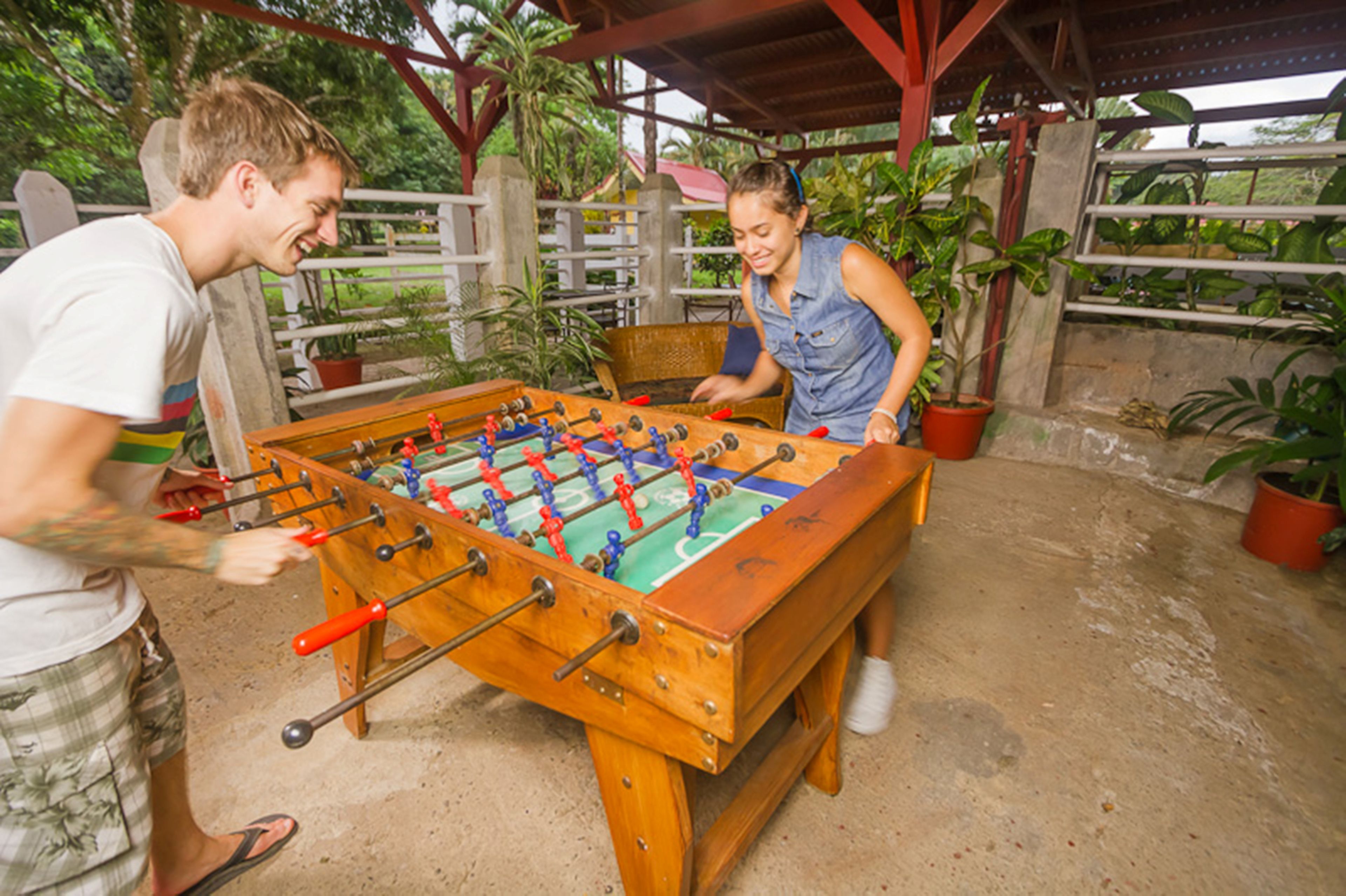 Uma mesa de futebol de madeira em ambiente externo, com piso de concreto e um cercado de madeira ao fundo.