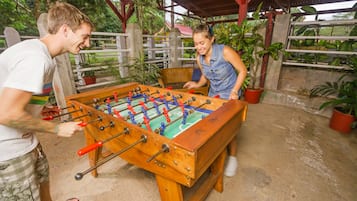 Uma mesa de futebol de madeira em ambiente externo, com piso de concreto e um cercado de madeira ao fundo.