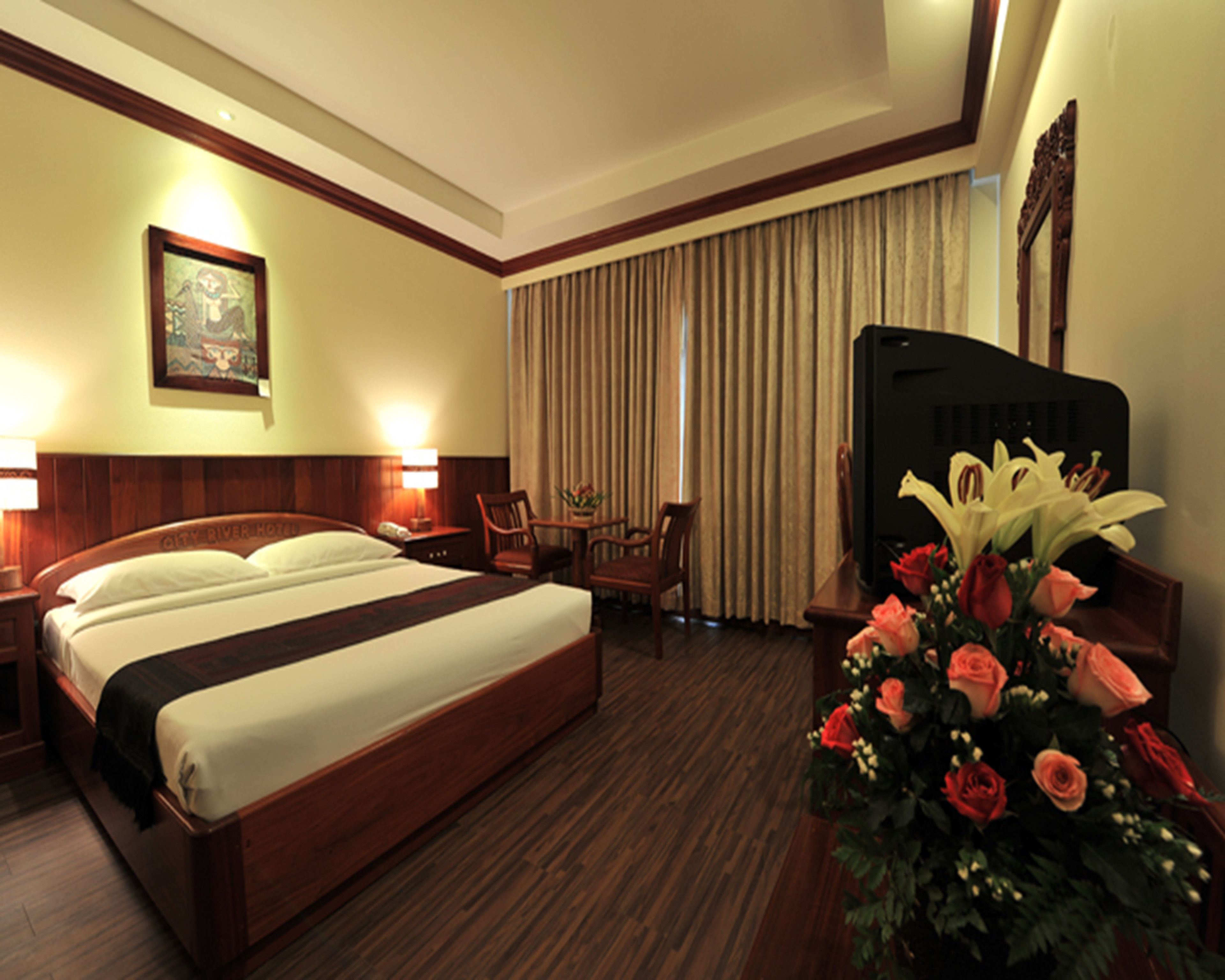 Quarto de hotel com uma cama grande, uma mesa com cadeira, uma televisão e um arranjo floral.