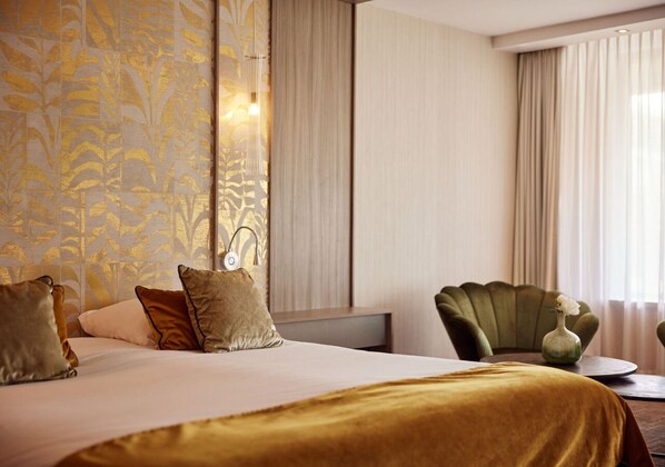 Quarto de hotel com uma cama, uma poltrona verde, uma mesinha redonda e um vaso com uma flor.