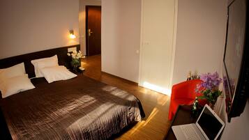 Quarto de hotel com uma cama, uma mesa com um laptop, uma cadeira, uma TV e uma porta.