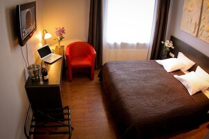 Quarto de hotel com cama, escrivaninha com laptop, cadeira, televisão e janela com cortinas.
