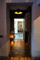 Um corredor com piso xadrez, luminária pendente e porta que dá para um quarto com uma cadeira e uma planta.