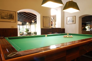 Uma mesa de bilhar com superfície em feltro verde, estrutura de madeira e iluminação pendente.