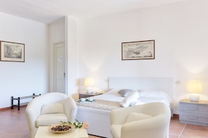 Quarto de hotel com cama, duas mesas de cabeceira, uma cadeira, uma mesinha com uma cesta de frutas e duas imagens emolduradas nas paredes.
