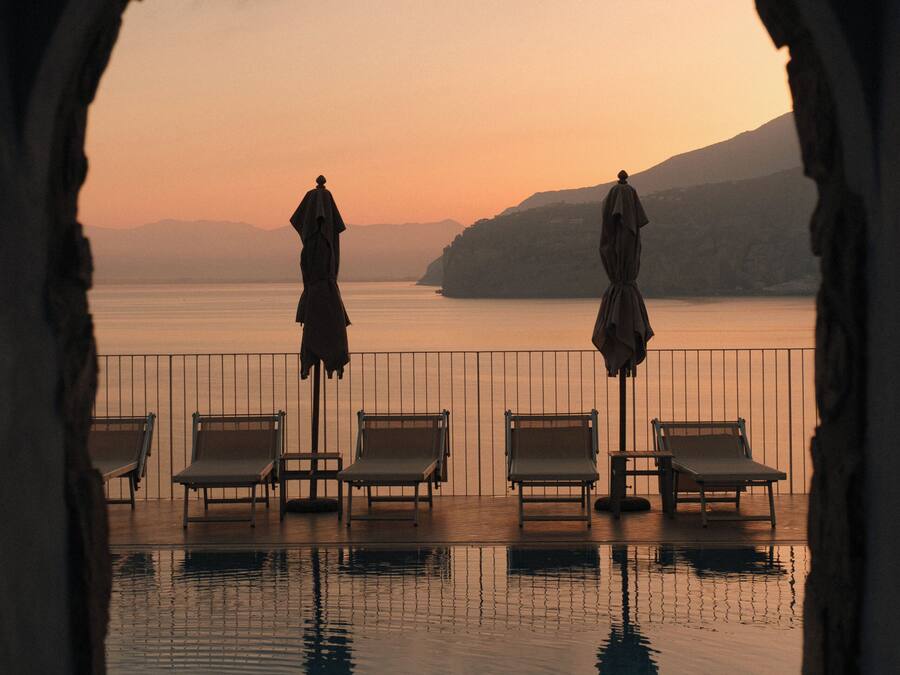 Uma piscina infinita com espreguiçadeiras e guarda-sóis, vista através de uma porta em arco, com vista para um corpo de água e terras distantes.