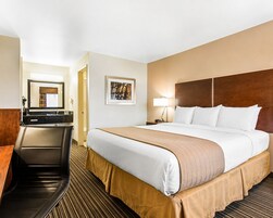 Quarto de hotel com cama, escrivaninha, cadeira, espelho, quadro e mesa de cabeceira com abajur.