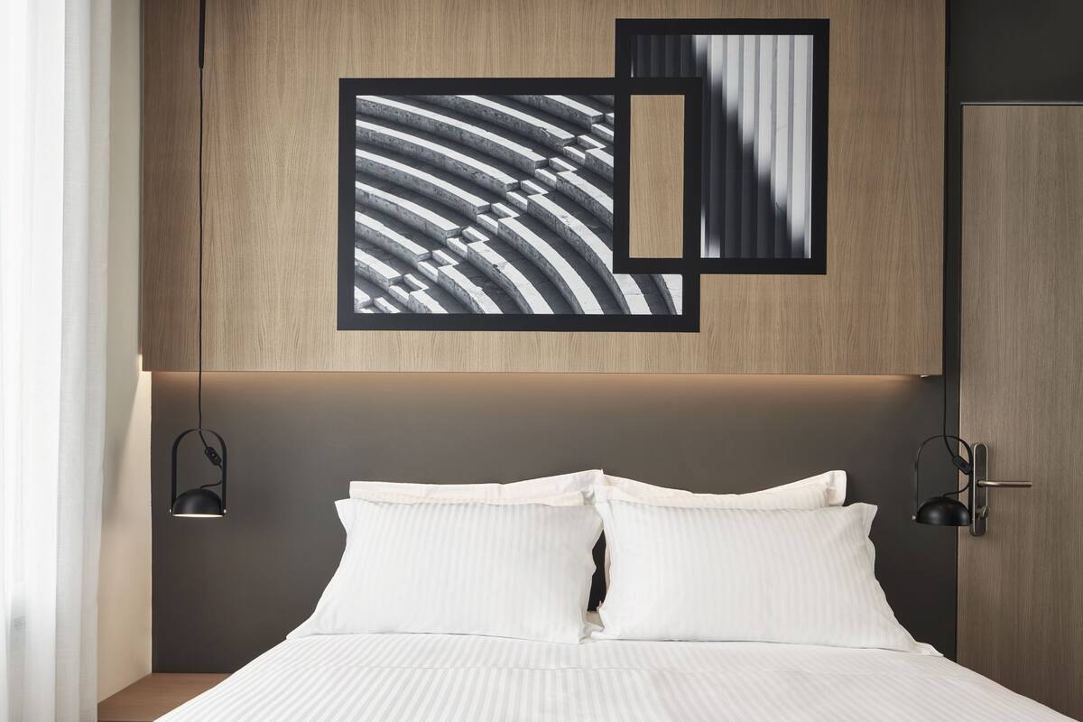 Quarto de hotel moderno com uma cama grande, duas luminárias laterais e uma obra de arte na parede.