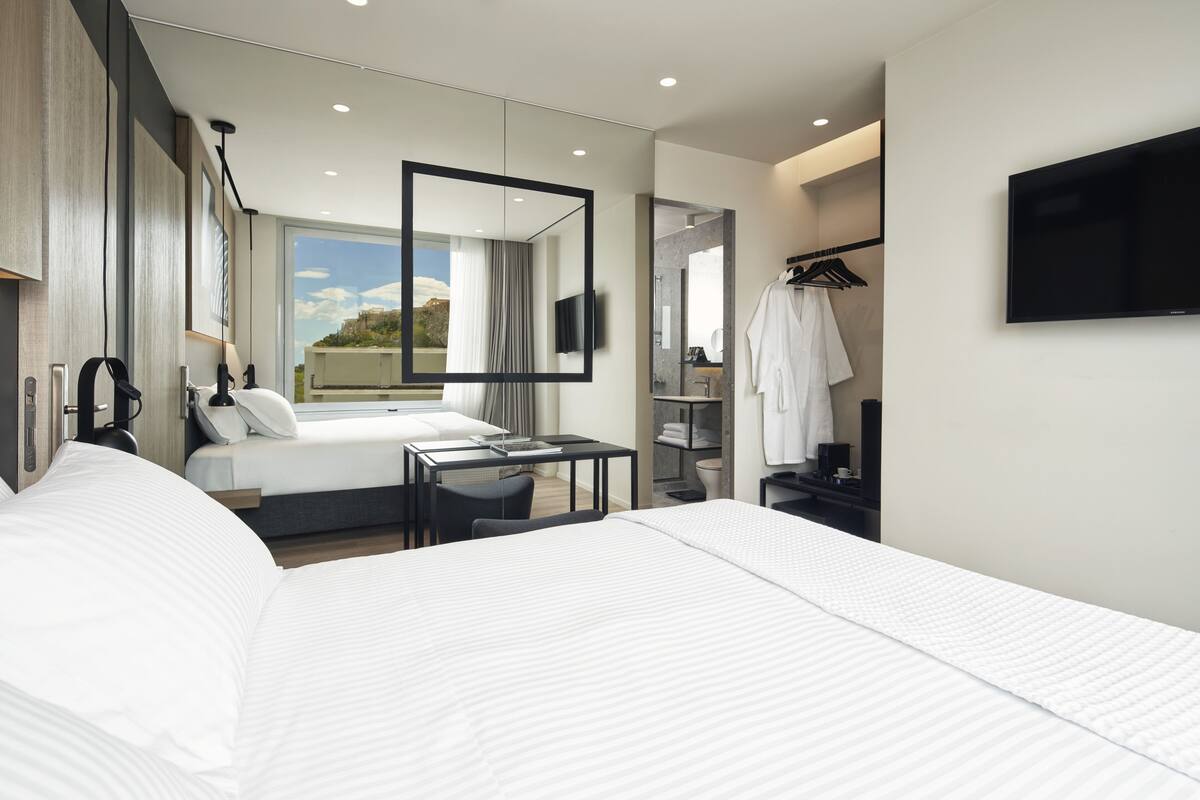 Quarto de hotel moderno com uma cama grande, uma escrivaninha, um espelho, uma televisão e vista para o exterior.
