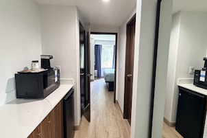 Um corredor estreito que dá para um quarto com cama e janela. Há duas máquinas de café pretas no balcão.