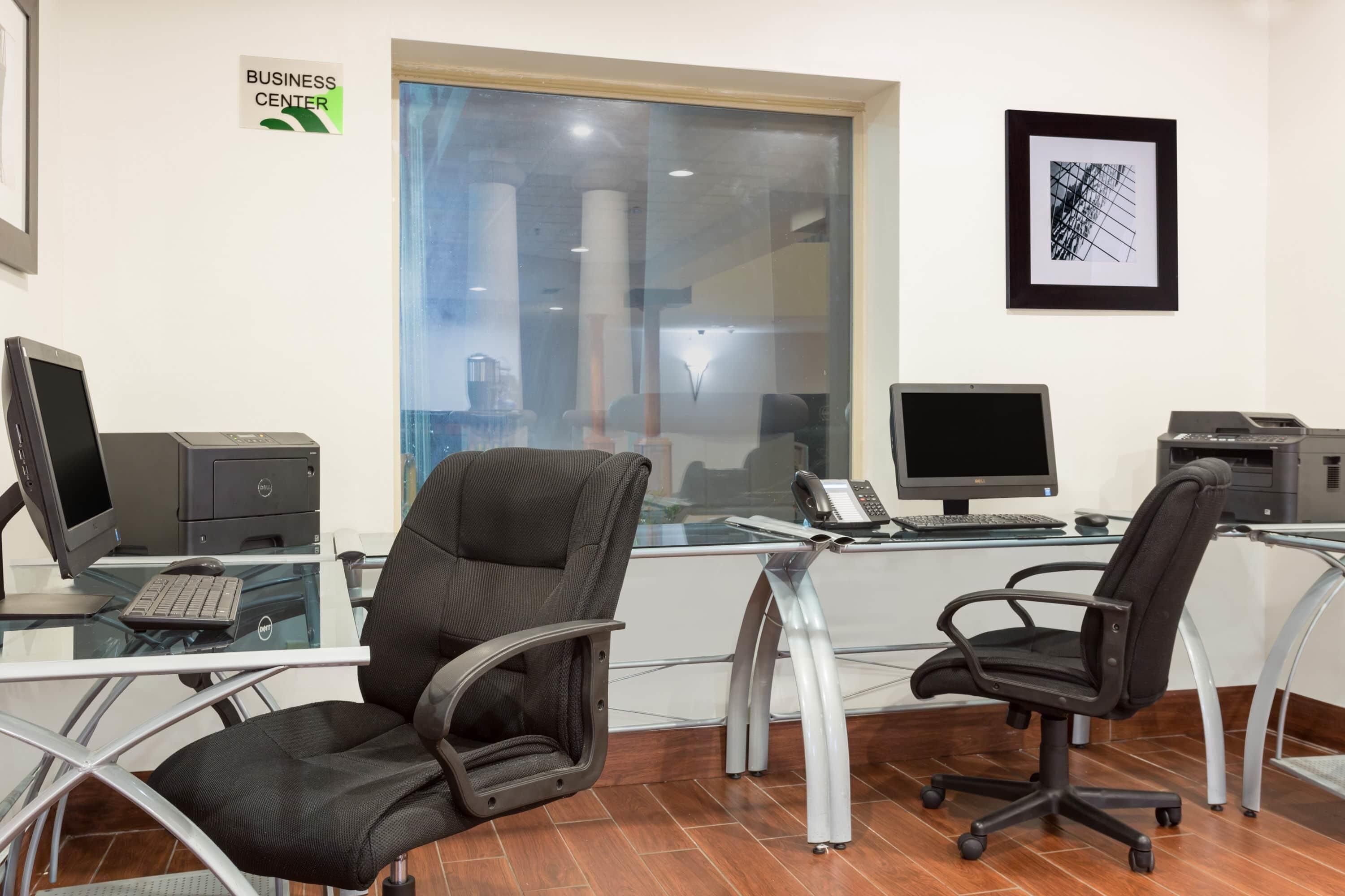 Um centro de negócios com mesas de vidro, cadeiras de escritório pretas, computadores e uma obra de arte abstrata emoldurada na parede.