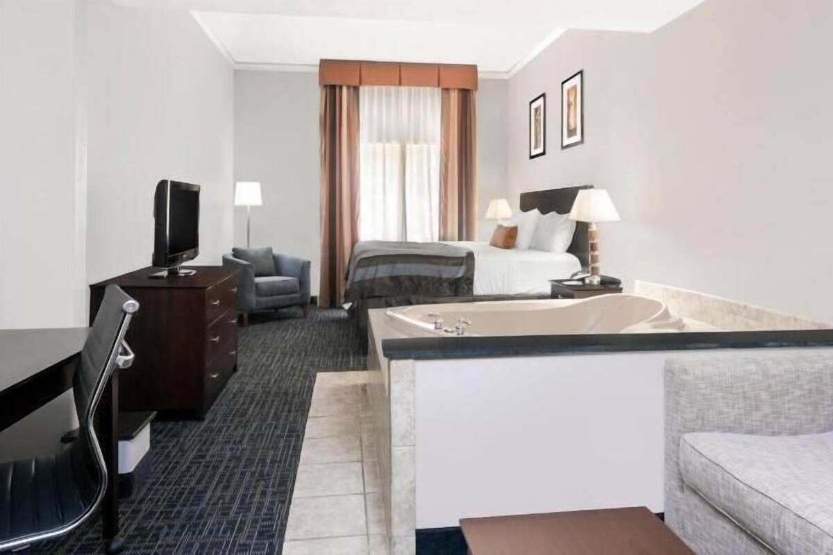 Quarto de hotel com cama, sofá, televisão, escrivaninha, penteadeira e banheiro com banheira.