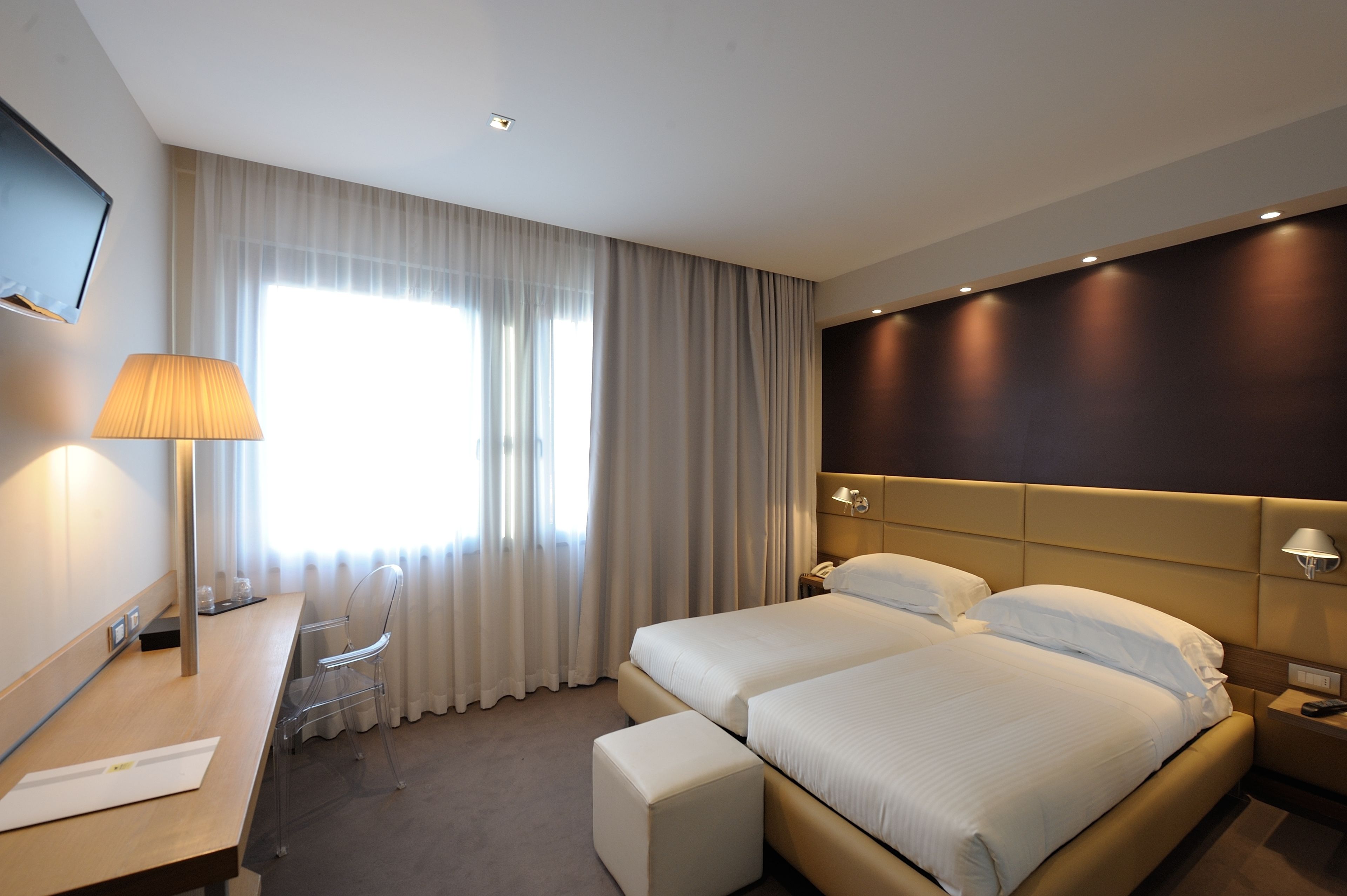 Quarto de hotel com duas camas, uma escrivaninha, uma cadeira, uma TV e uma janela com cortinas.