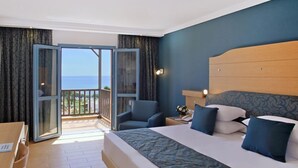 Quarto de hotel com uma cama grande, cabeceira de madeira, uma escrivaninha e varanda com vista para o mar.