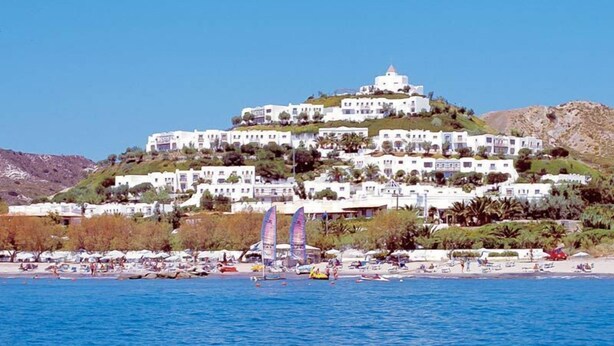 Uma vila costeira com edifícios brancos numa encosta, uma praia em primeiro plano e águas azul-claras.
