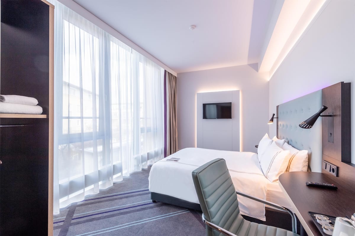 Quarto de hotel com cama, escrivaninha, cadeira, televisão e uma janela grande com cortinas.