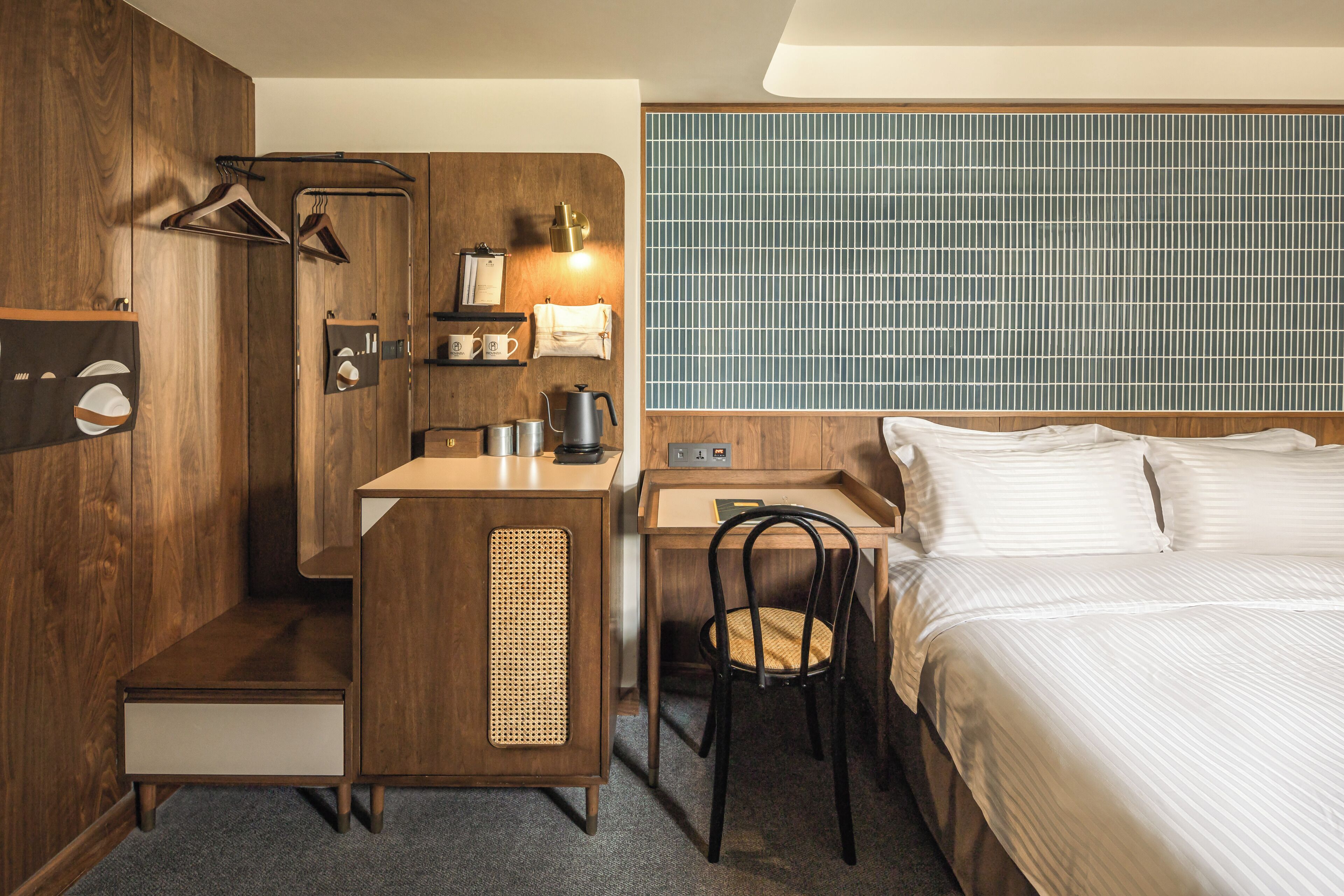 Quarto de hotel com guarda-roupa de madeira, uma escrivaninha com cadeira, uma cama e parede com padrão quadriculado azul.