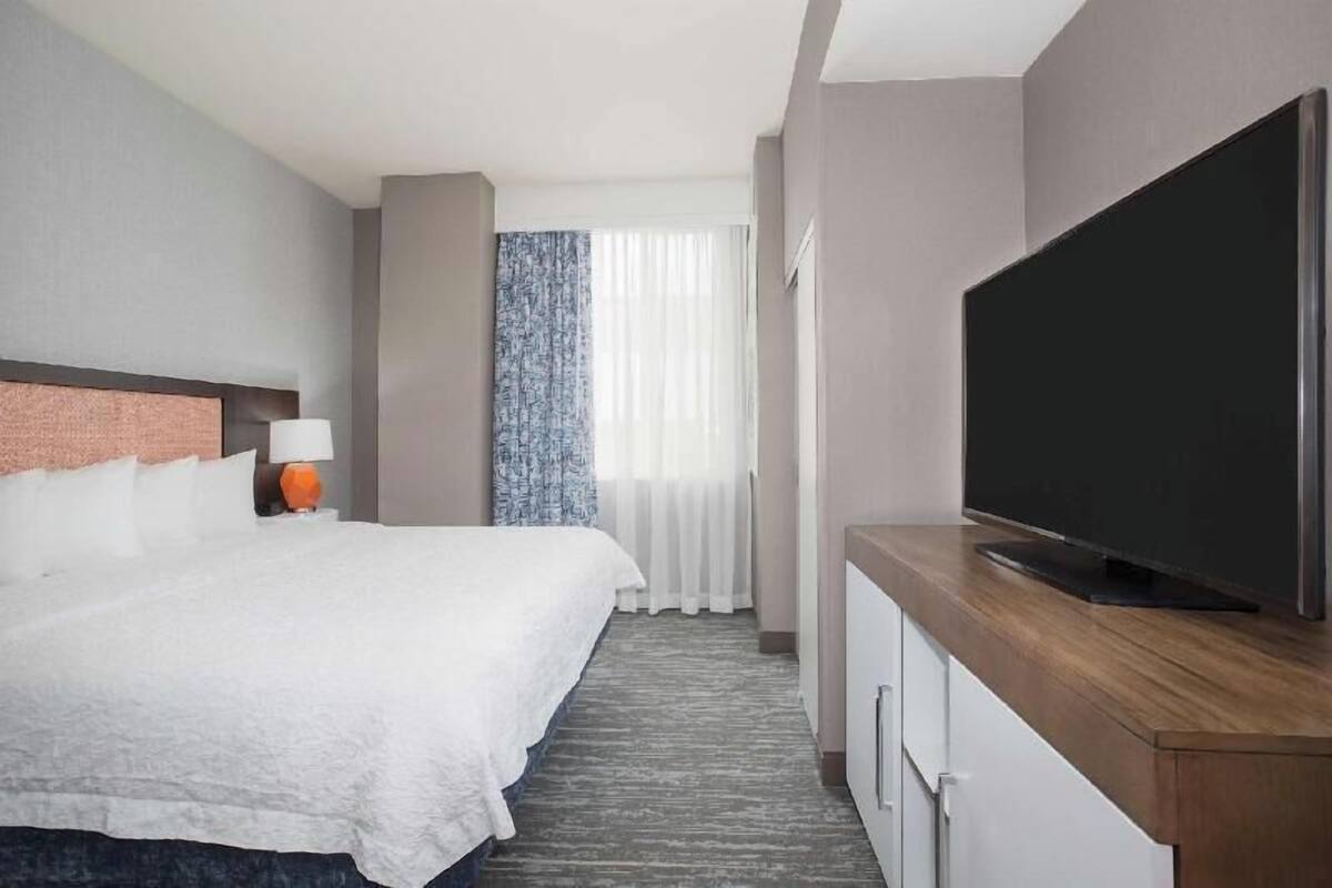 Quarto de hotel com cama, televisão sobre um móvel de madeira, janela com cortinas e luminária fixada na parede.