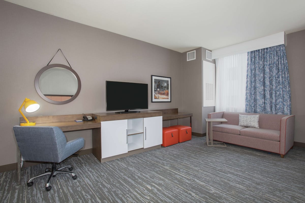 Quarto de hotel com escrivaninha, cadeira, televisão, sofá e uma janela grande com cortinas.