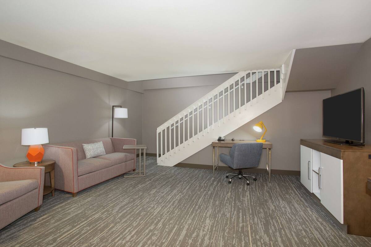 Quarto de hotel com escada, sofá, escrivaninha, televisão e luminária.