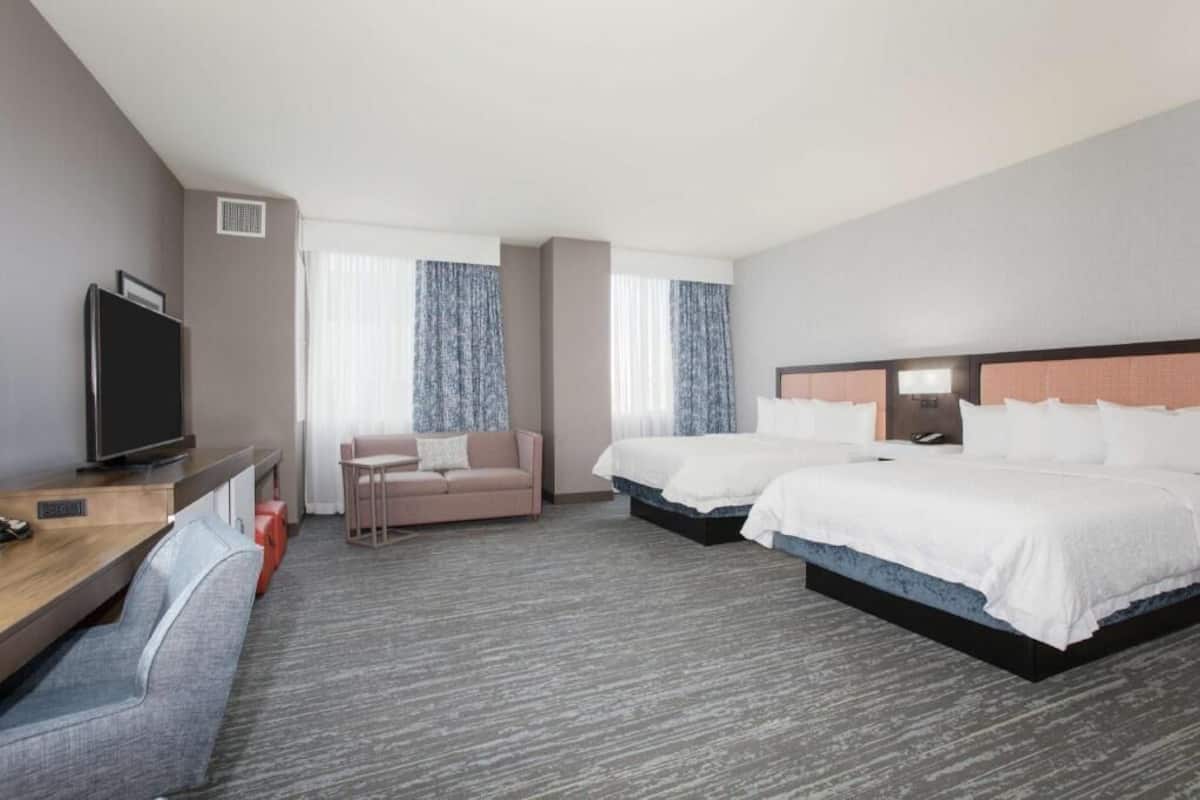 Quarto de hotel com cama, escrivaninha, televisão, sofá e janela com cortinas.