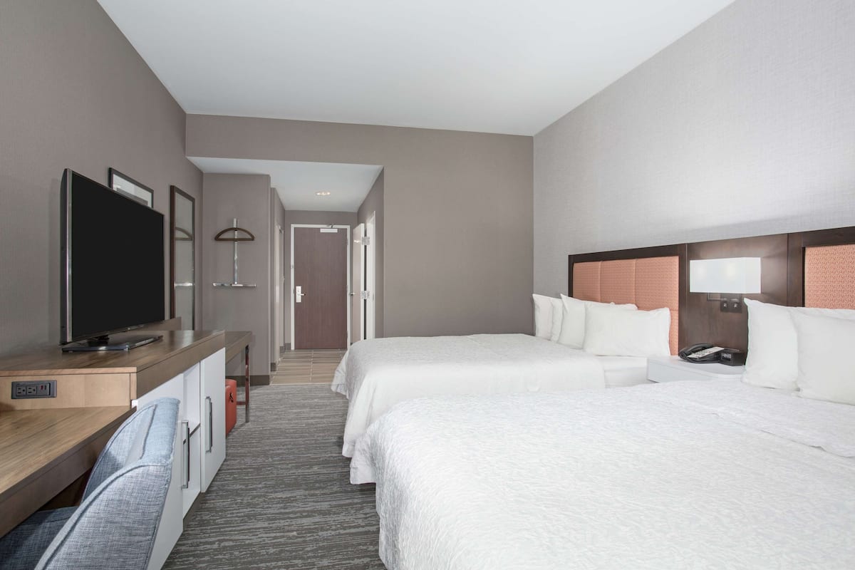 Quarto de hotel com duas camas, uma escrivaninha com televisão, uma lâmpada e um telefone.