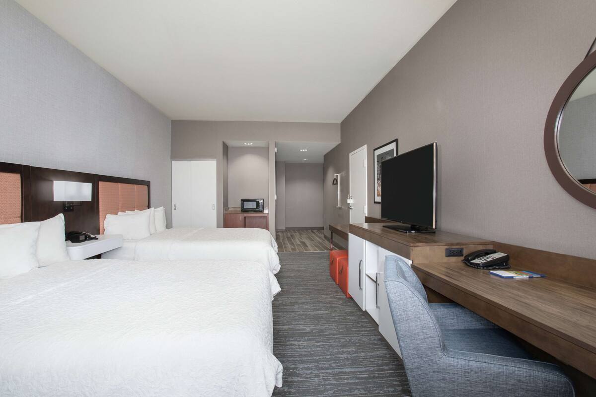 Quarto de hotel com duas camas, uma escrivaninha com computador, televisão e banheiro ao fundo.