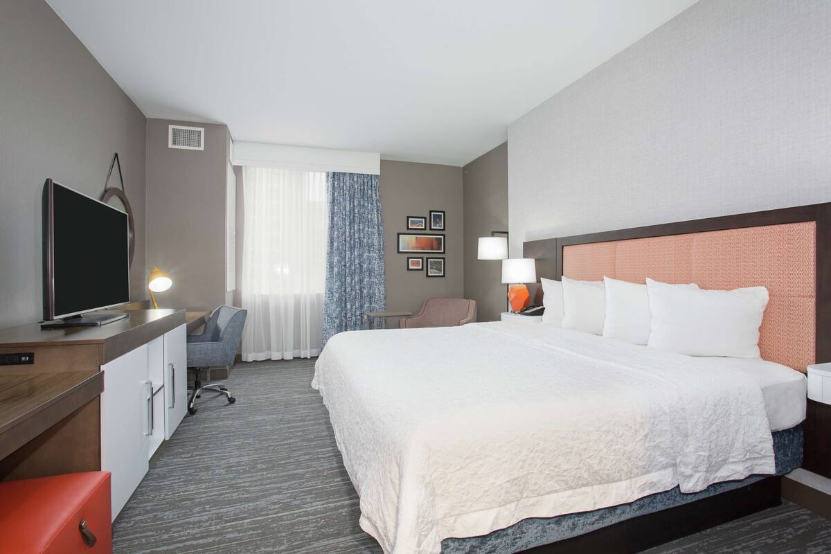 Quarto de hotel com uma cama grande, uma escrivaninha com televisão, uma cadeira, luminária de mesa e janela com cortinas.
