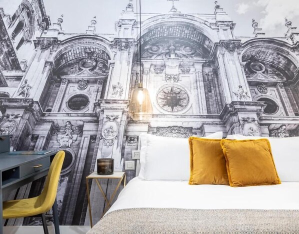 Quarto com um grande mural de um edifício histórico, cama com almofadas brancas e mostarda, escrivaninha com cadeira e uma pequena mesa com uma vela.