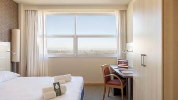 Quarto de hotel com cama, escrivaninha e vista para a paisagem urbana pela janela.