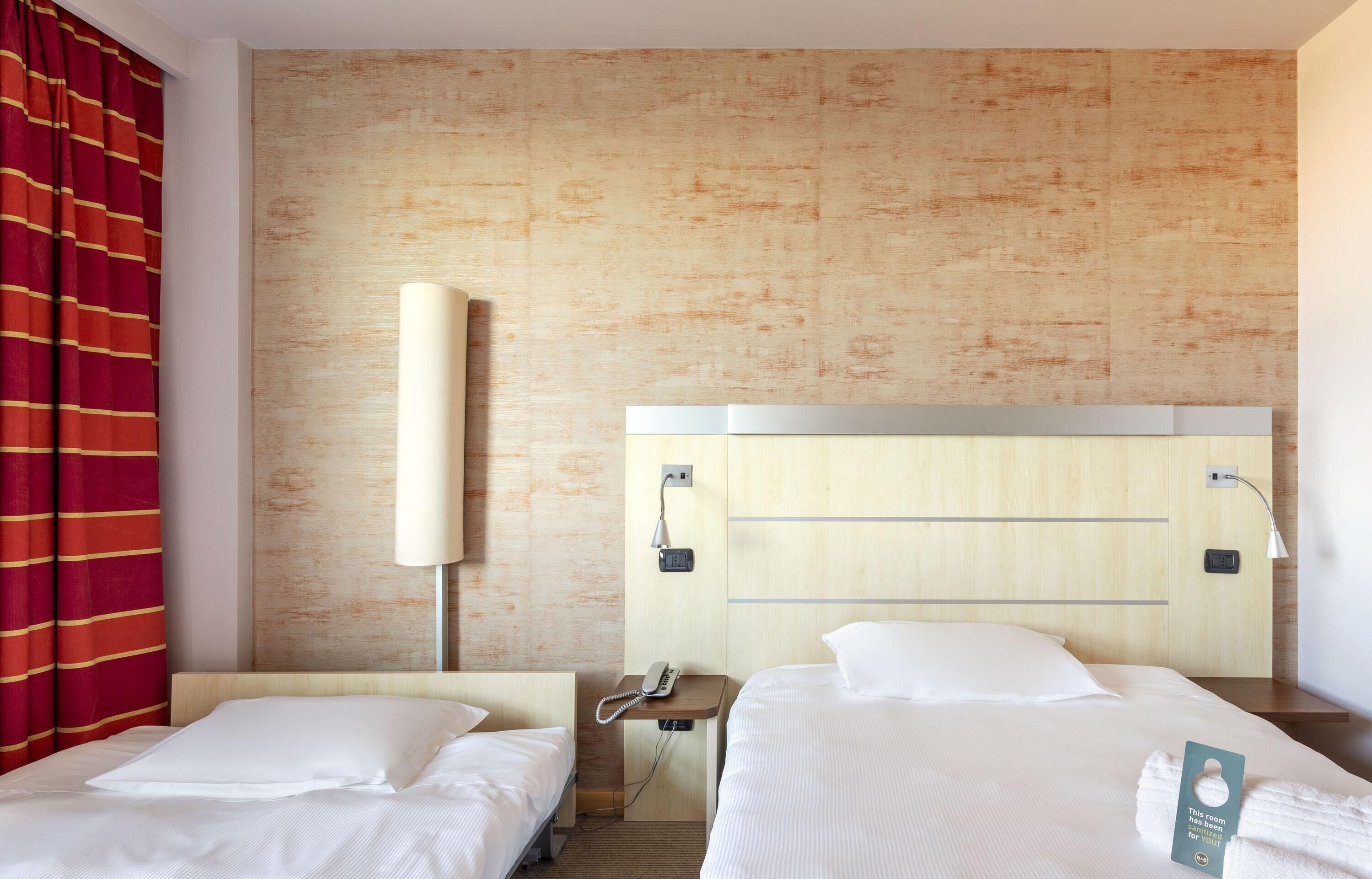 Quarto de hotel com duas camas, cabeceira de madeira, mesa de cabeceira com abajur, telefone e cortina vermelha.
