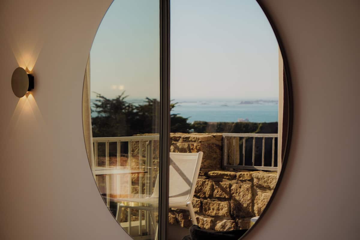 Janela circular com vista para uma parede de pedra, uma cadeira branca e uma mesa.