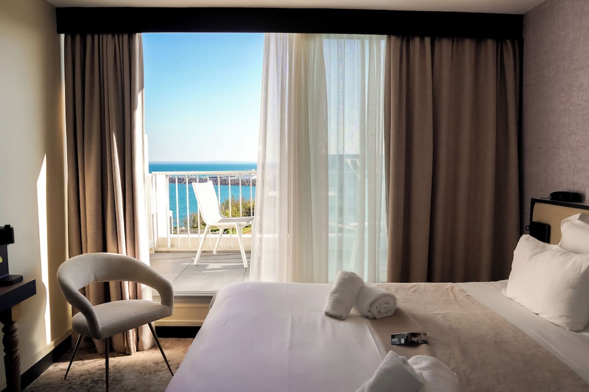 Quarto de hotel com cama, cadeira, escrivaninha e vista para o mar.