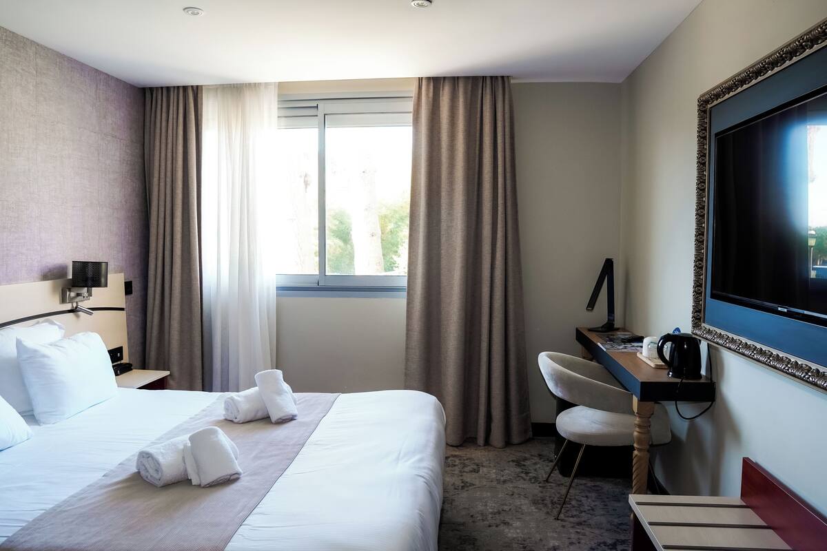 Quarto de hotel com cama, escrivaninha, cadeira, televisão e janela com cortinas.
