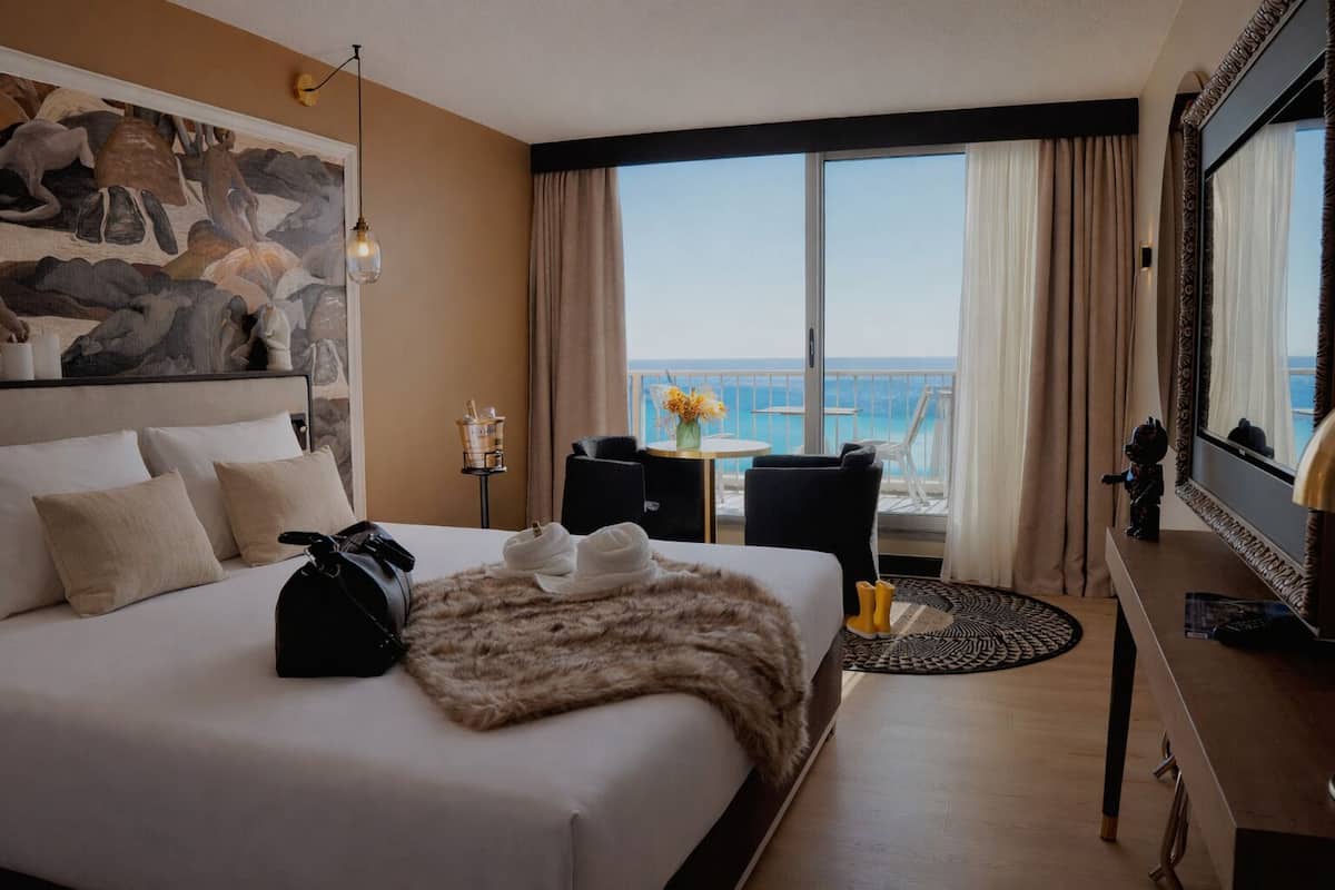 Quarto de hotel com cama grande, escrivaninha, cadeira, vista para o mar e um quadro na parede.