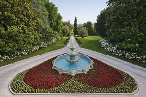 Um jardim simétrico com uma fonte central, rodeado por canteiros de flores bem cuidados e árvores altas.