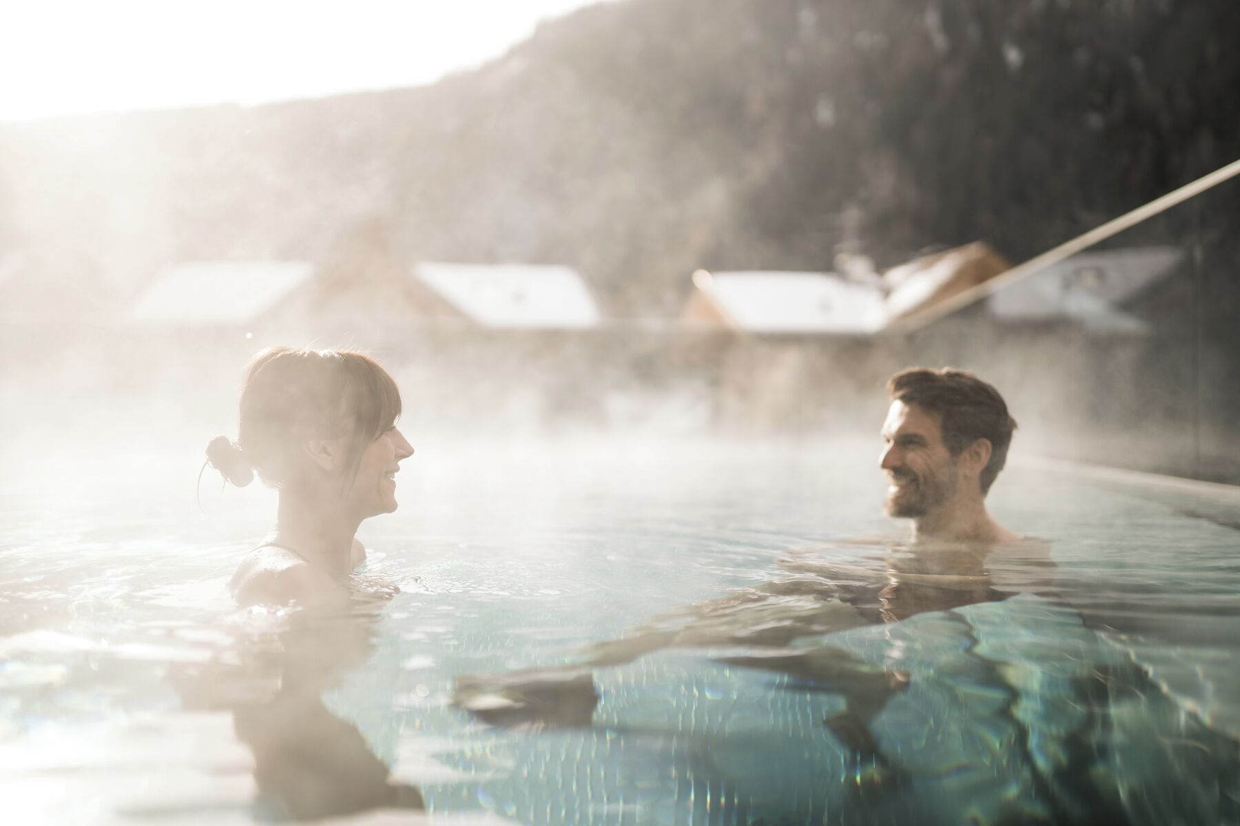 Um casal em uma piscina transparente com montanhas ao fundo.