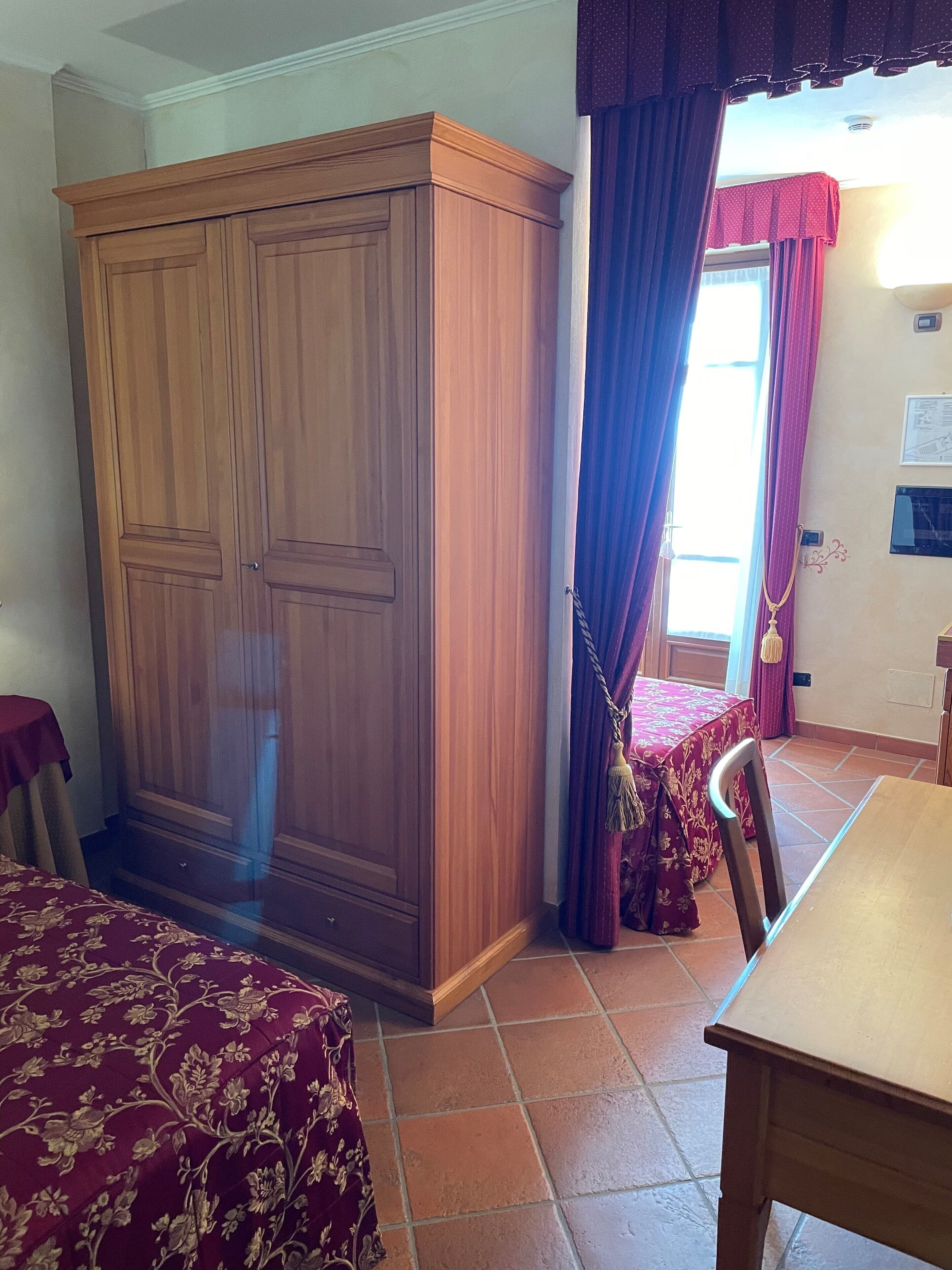 Quarto com armário de madeira, cama com colcha florida, mesa de jantar e janela com cortinas.