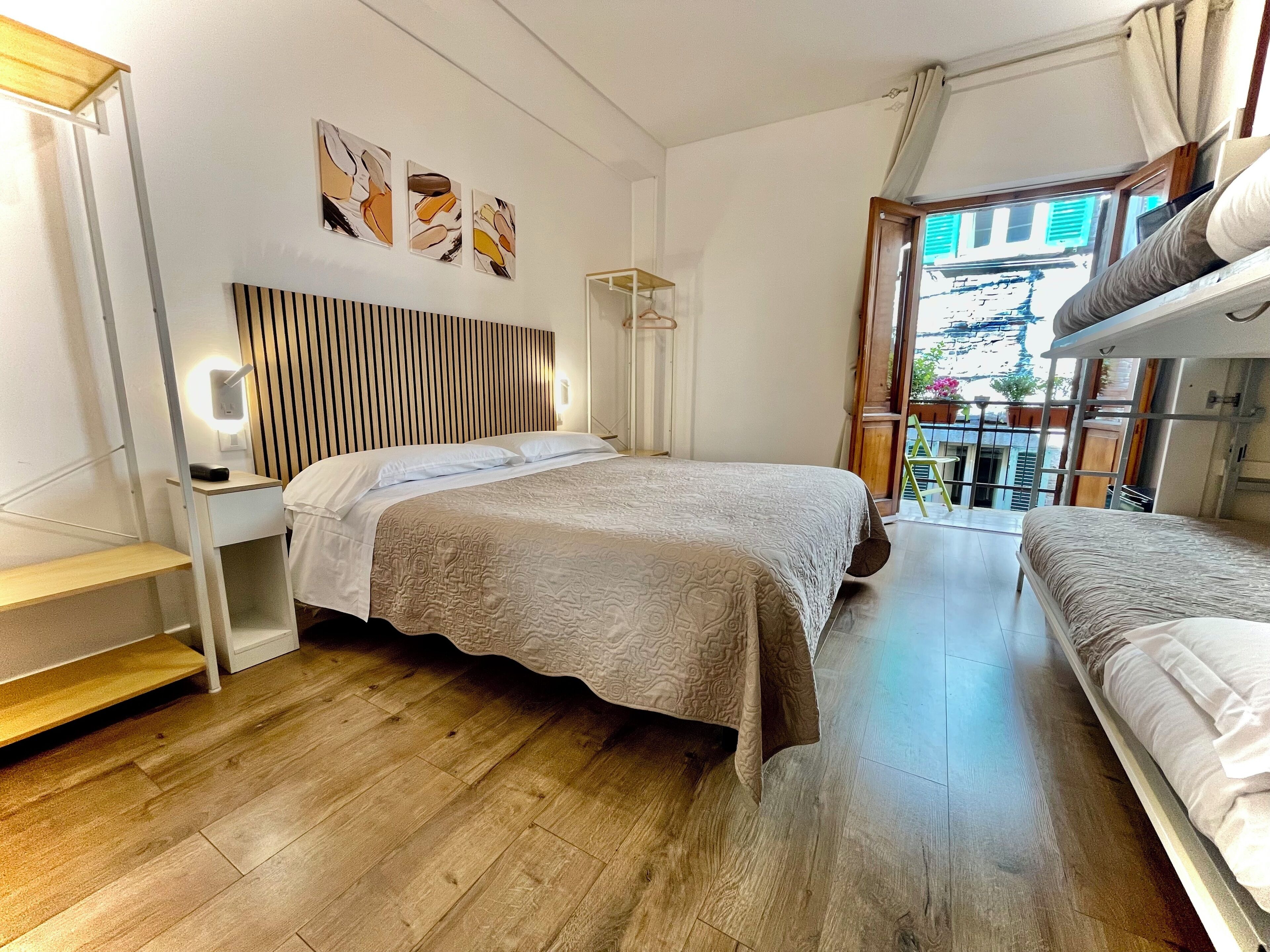 Quarto de hotel com uma cama grande, uma mesinha de cabeceira, uma luminária na parede e uma janela com vista para o exterior.