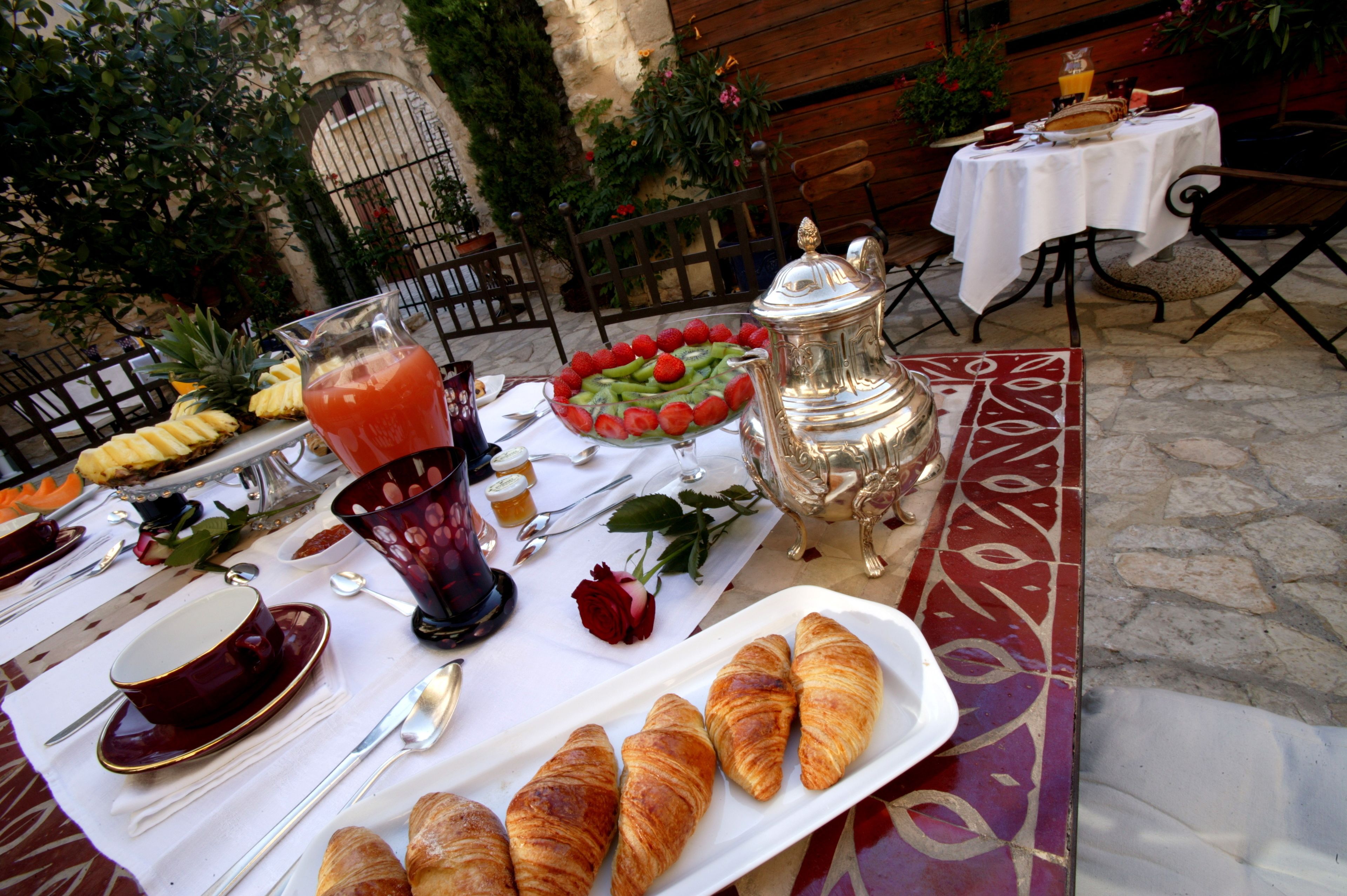 Uma área de refeições ao ar livre com uma mesa posta para uma refeição, incluindo croissants, uma tábua de frutas e uma jarra de suco.