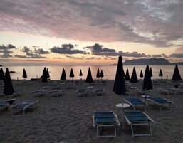 Uma praia com fileiras de espreguiçadeiras e guarda-sóis, mar calmo e céu ao pôr do sol.