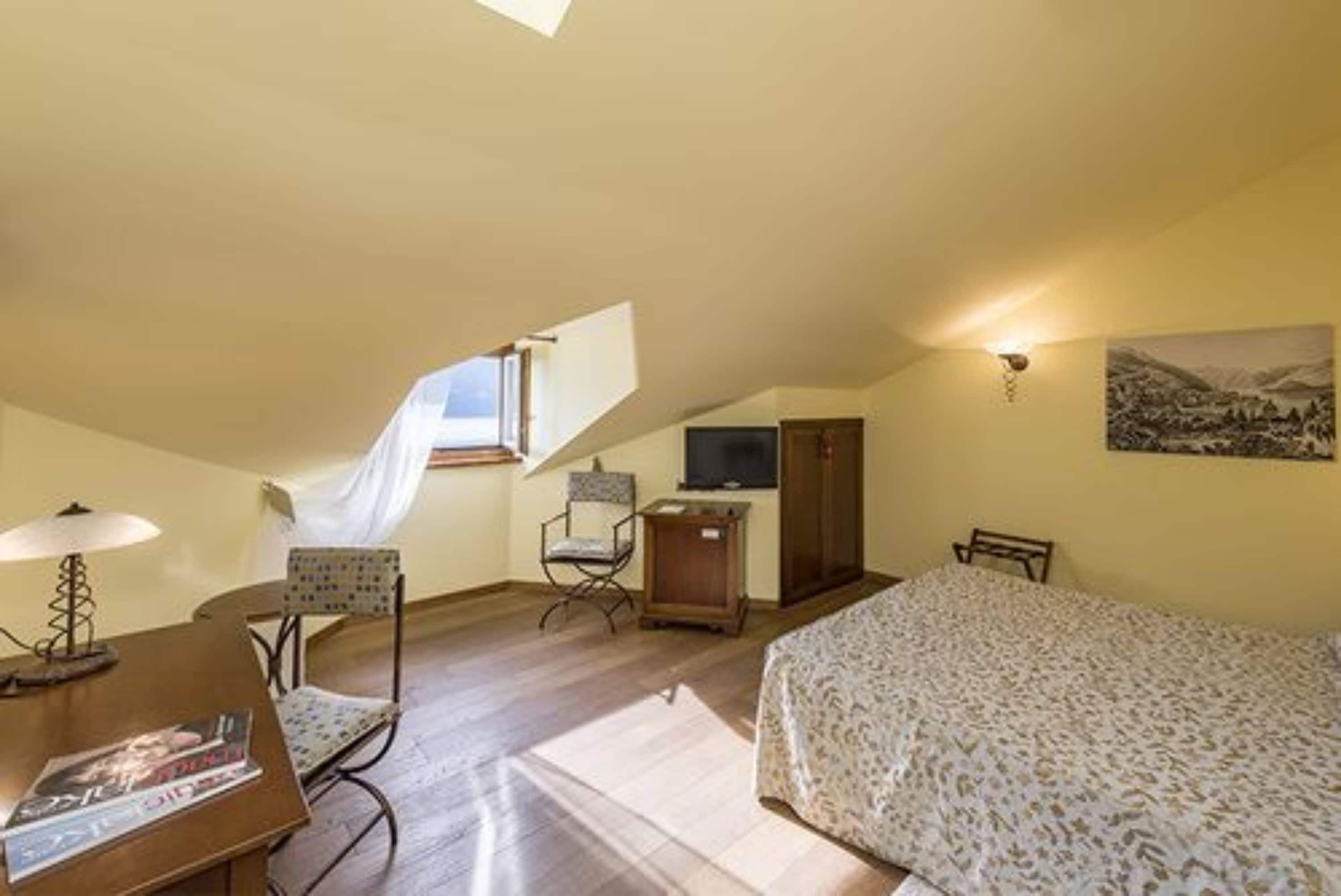 Quarto com cama, escrivaninha, cadeira, mesa pequena, janela e televisão fixada na parede.