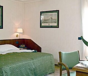 Quarto de hotel com cama, uma cadeira, escrivaninha, abajur e um quadro na parede.