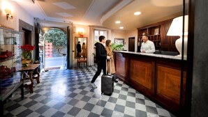 Um saguão de hotel com piso quadriculado, balcão de recepção de madeira e uma vitrine de vidro com objetos vermelhos.