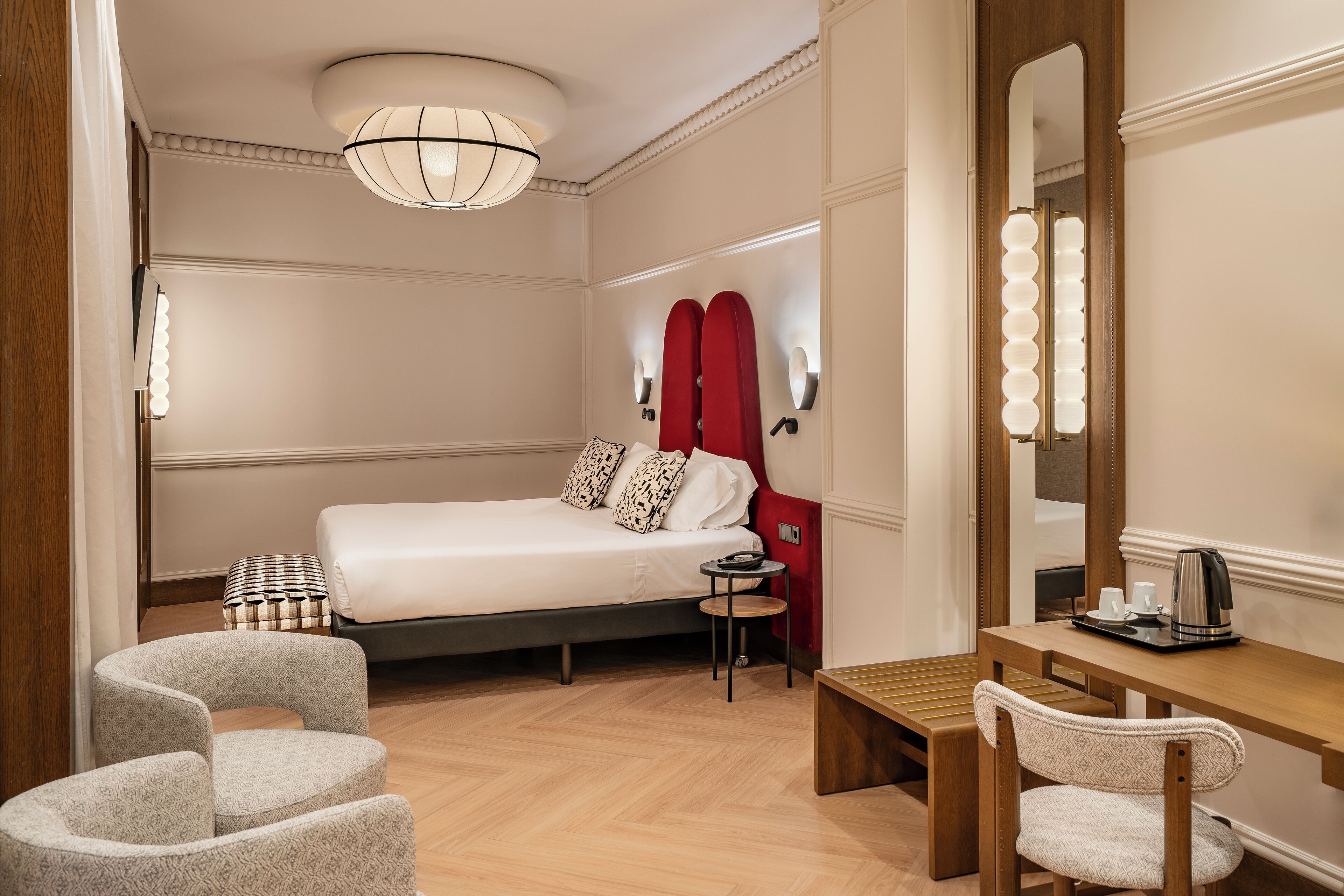 Quarto de hotel com uma cama, duas poltronas, uma secretária de madeira, um espelho iluminado e uma mesinha com chaleira.