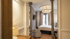 Quarto de hotel com piso de madeira, uma cama, uma cadeira, uma mesa e um espelho grande.