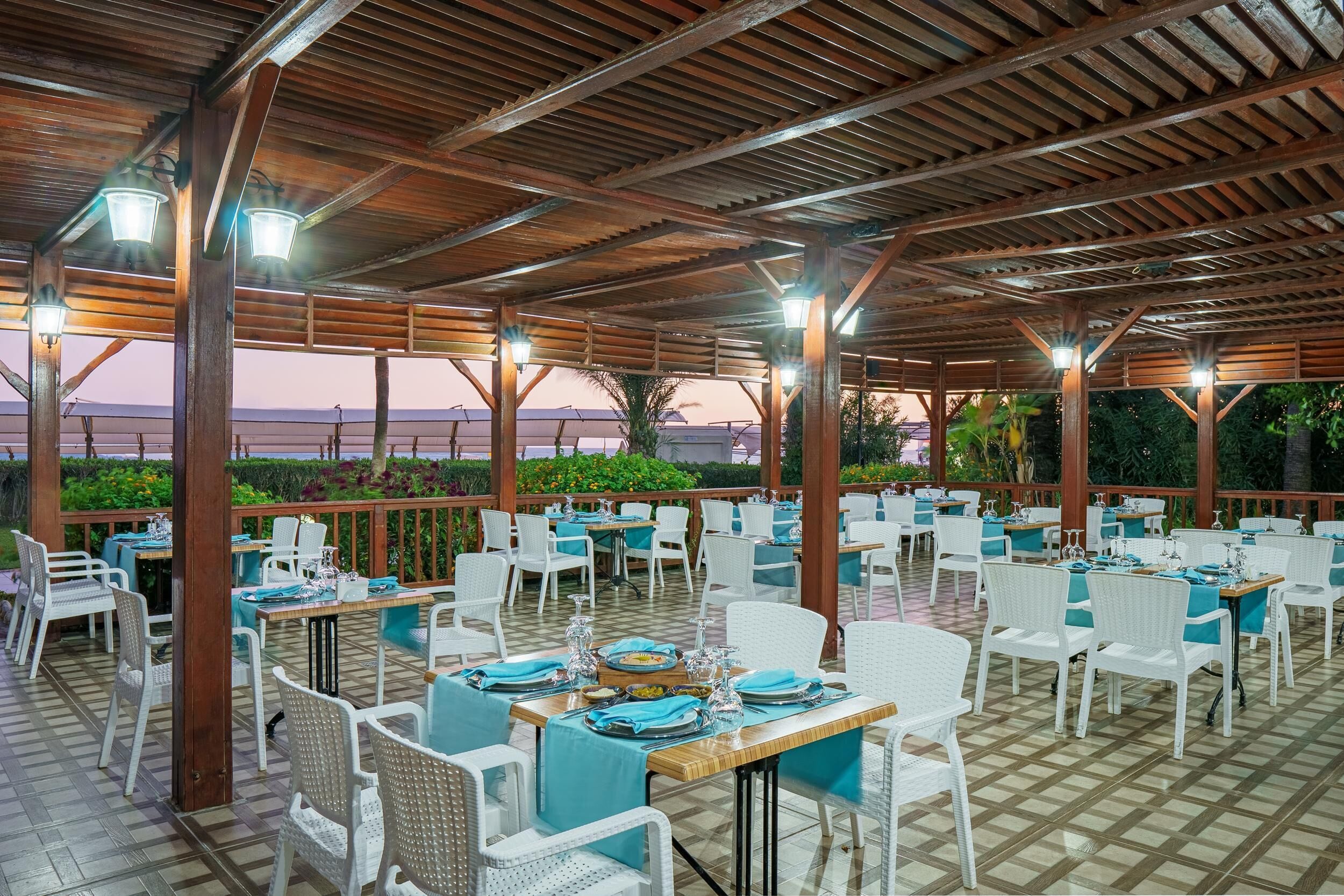 Um restaurante ao ar livre com vigas de madeira, cadeiras de vime branco e mesas preparadas para refeições.